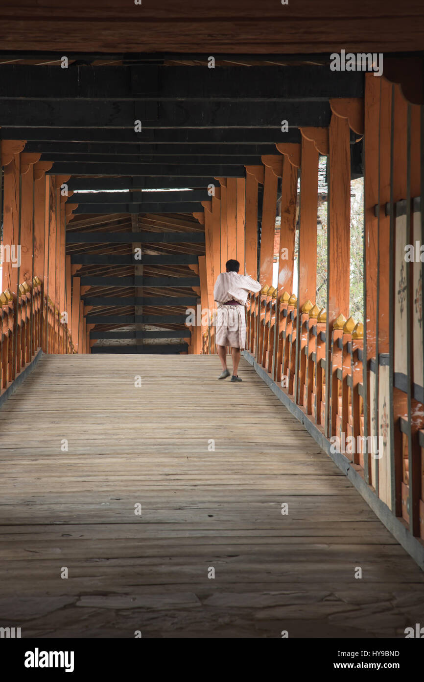 A Bhutanese man in the traditional gho robe crosses the covered bridge ...