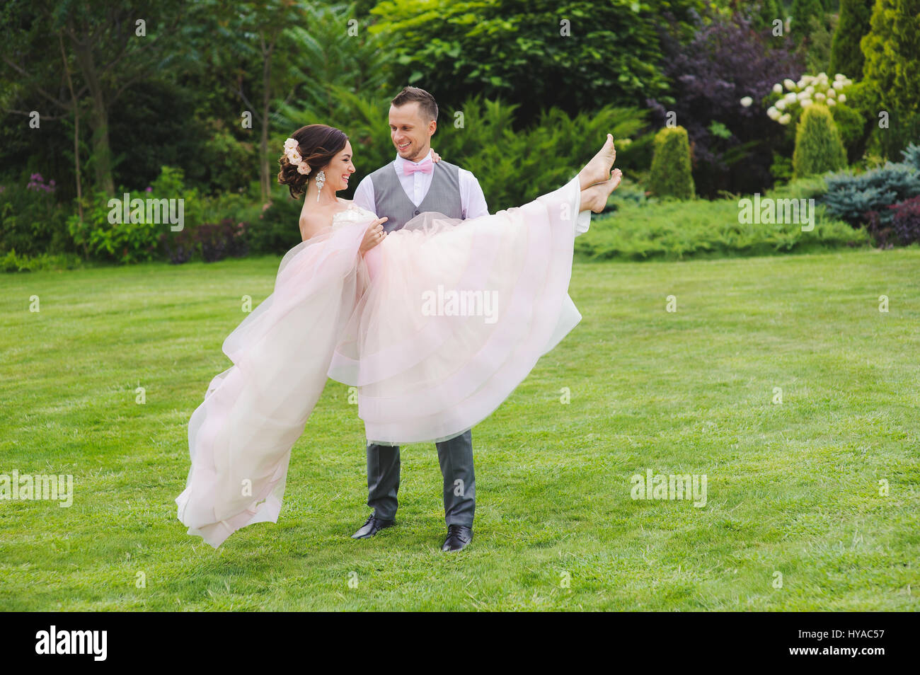 Groom holding beautiful bride in his arms outdoors. Pink wedding dress ...