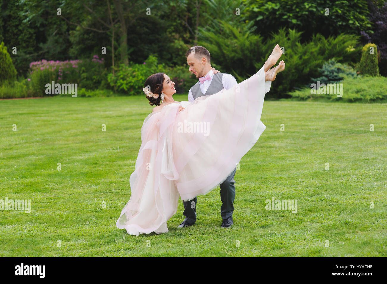 Groom holding beautiful bride in his arms outdoors. Pink wedding dress ...