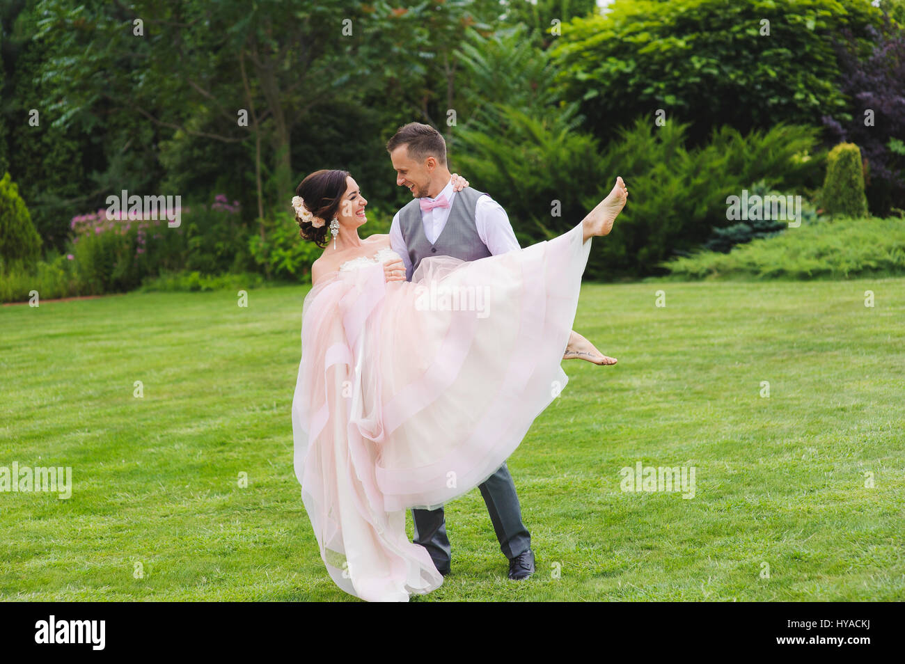 Groom holding beautiful bride in his arms outdoors. Pink wedding dress ...