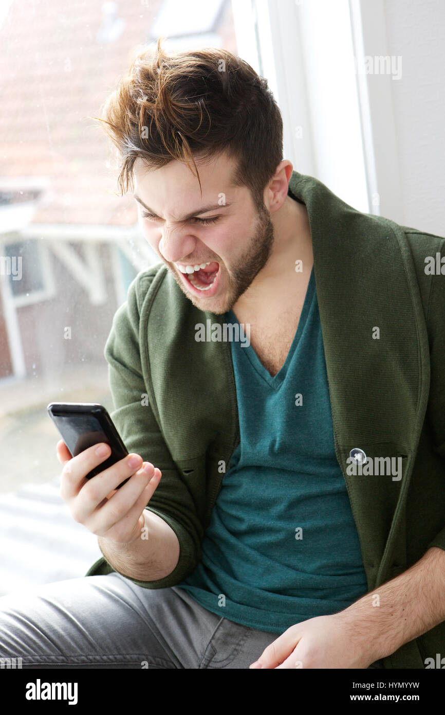 Close up portrait of a young man looking at mobile phone with angry ...