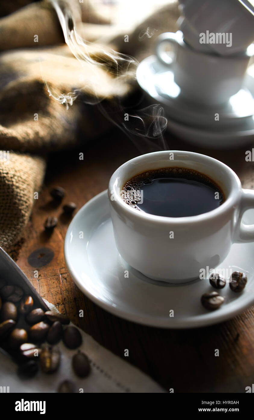 Cup of espresso steaming with coffee beans in foreground and mugs in ...