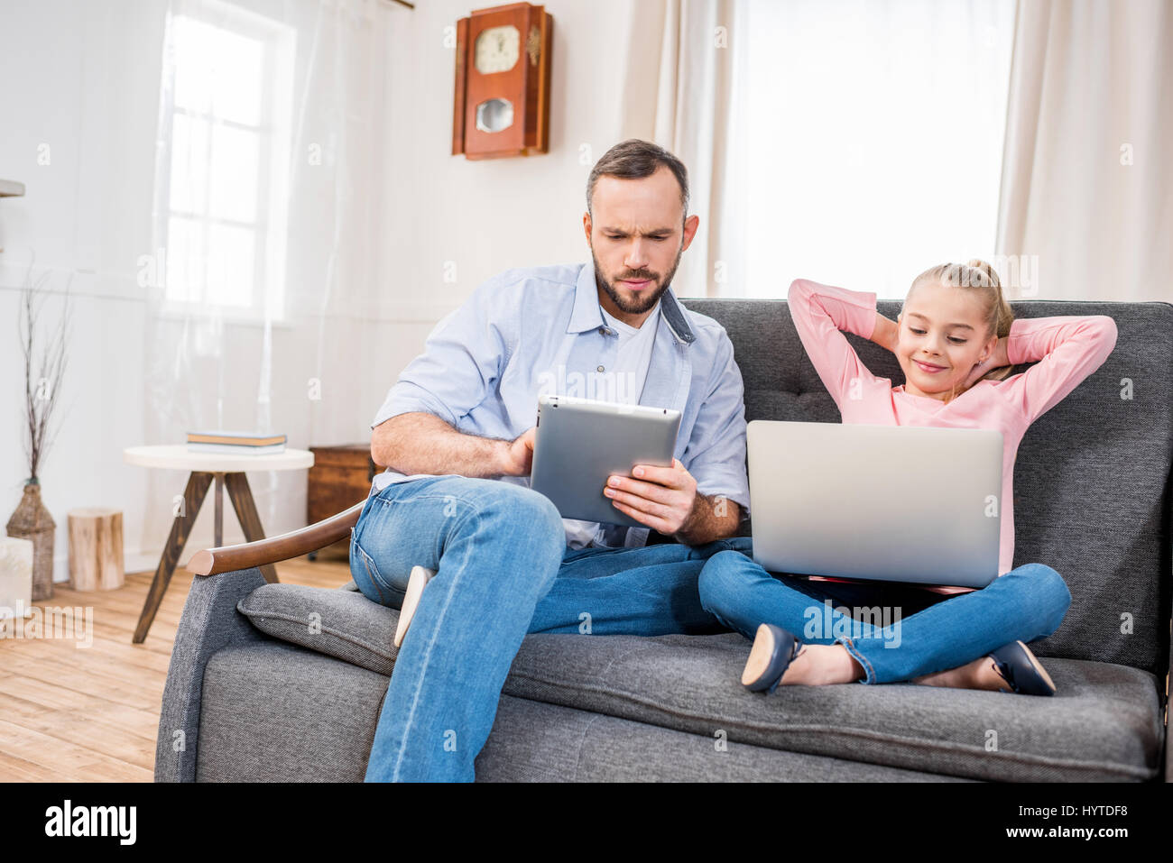 Serious father and cute smiling daughter using digital devices on couch ...
