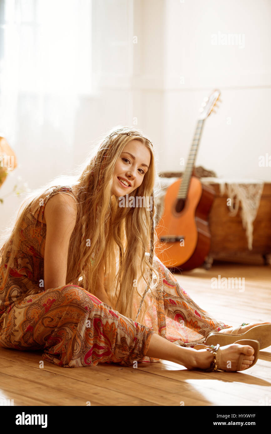 Smiling young hippie woman with long hair sitting on floor and looking ...