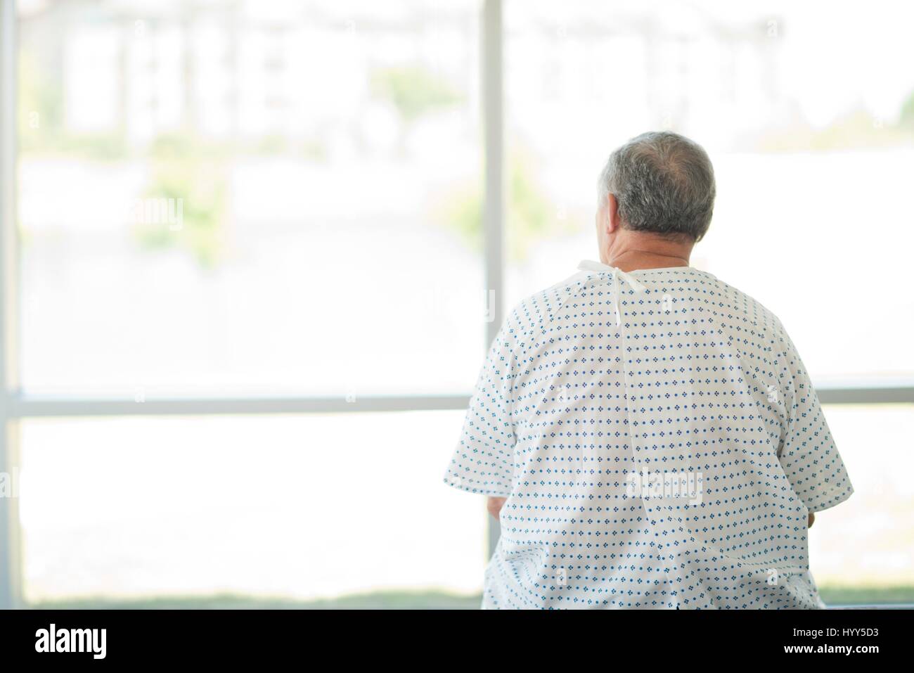Male patient wearing gown looking through hospital window, rear view ...