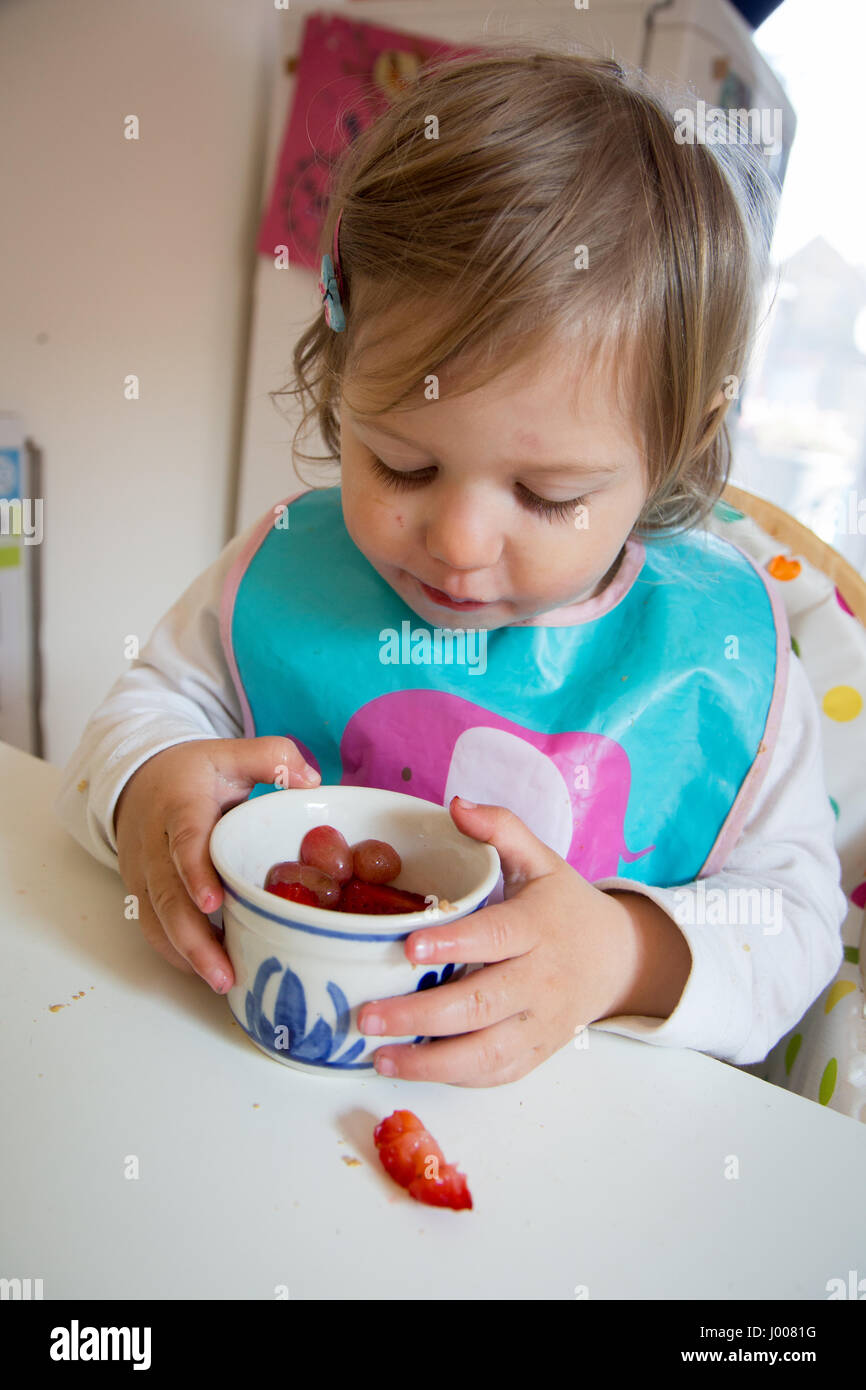 A girl toddler eating strawberries enthusiastically at a table. She is ...