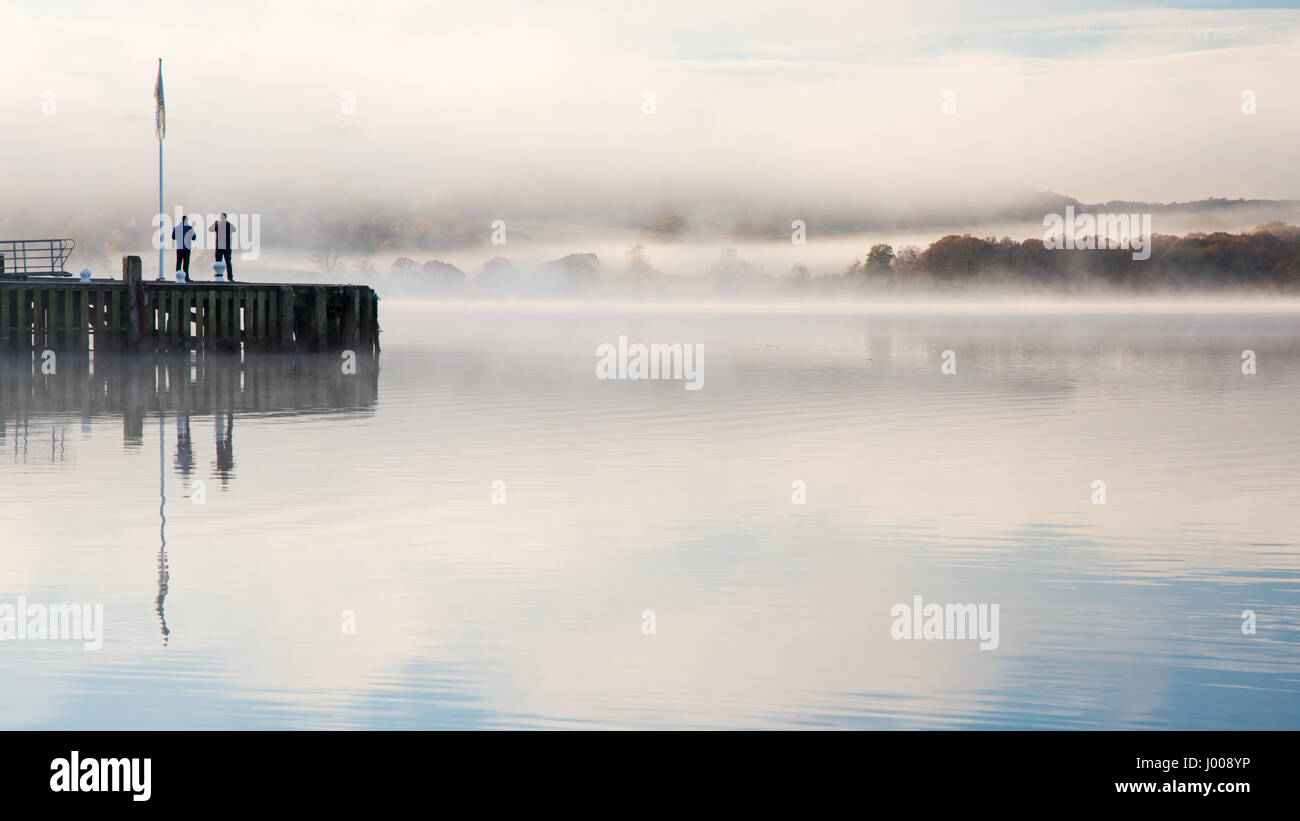 Two people stand on Waterhead Pier in a misty Windermere lake at ...