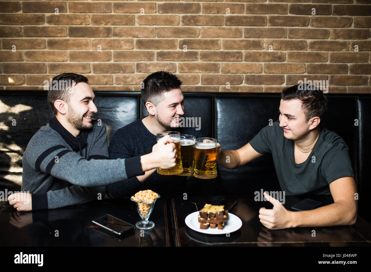 Three happy young men in casual wear talking and drinking beer while ...