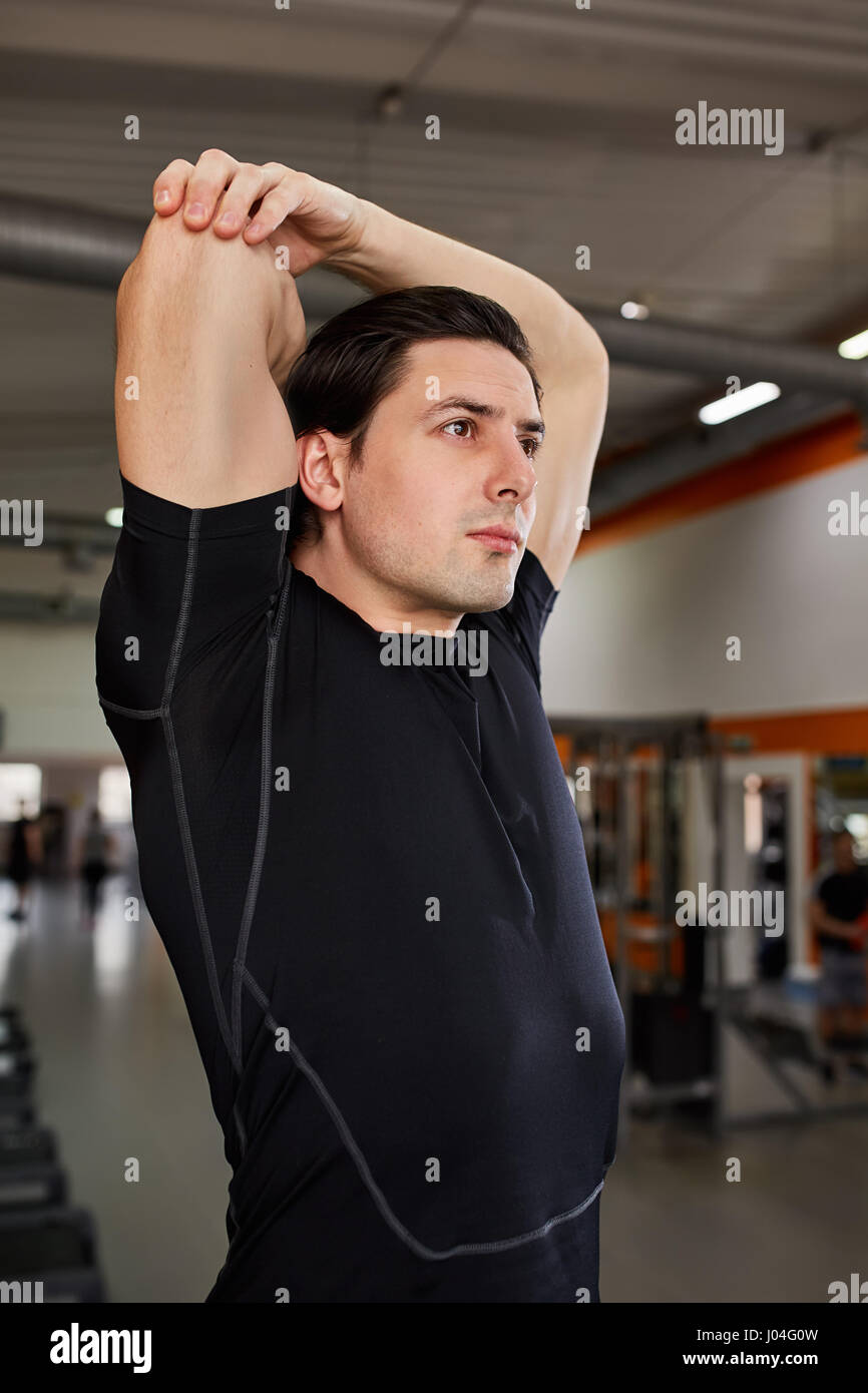 Full-length of young man in black sportswear stretching his arm at gym ...