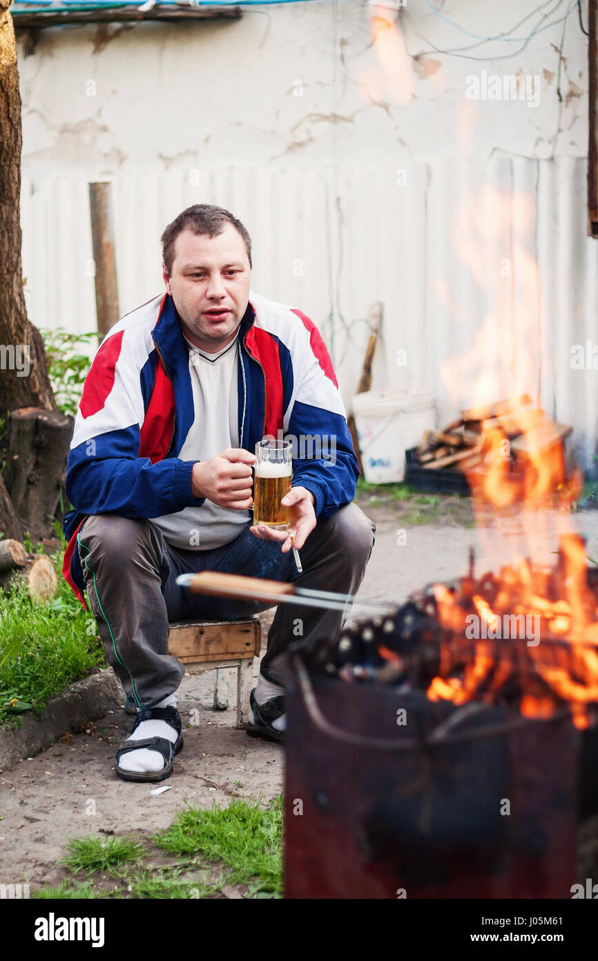 A young adult man smokes and drinks beer near the barbecue with a fire ...