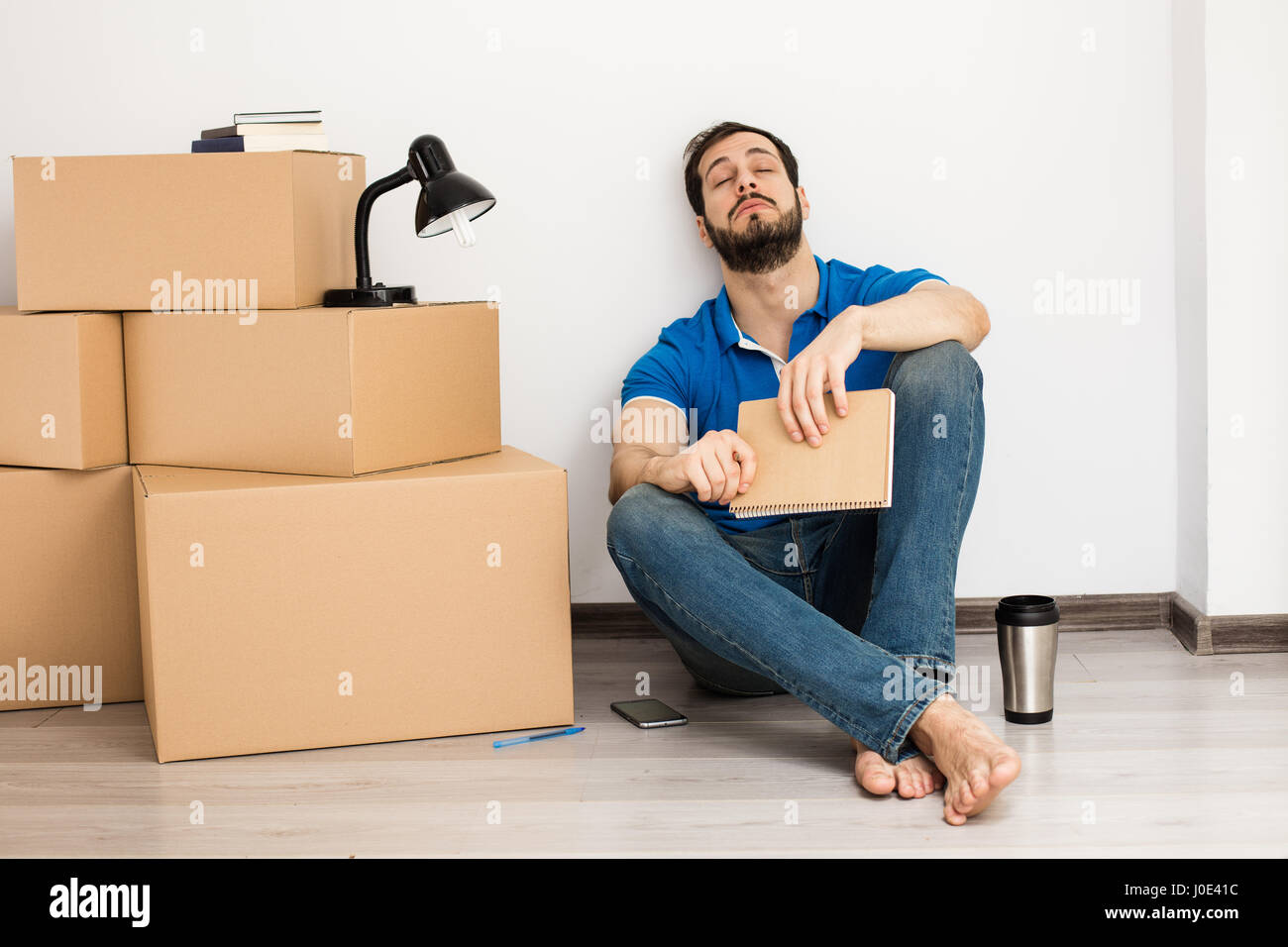 tired young man lying on the floor with packing boxes near and coffee ...