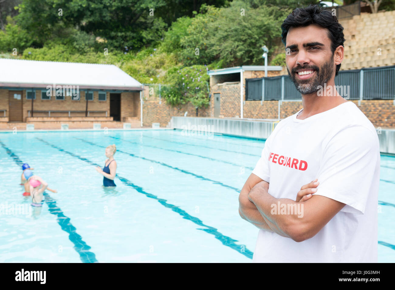Portrait of smiling lifeguard standing with arms crossed in poolside ...