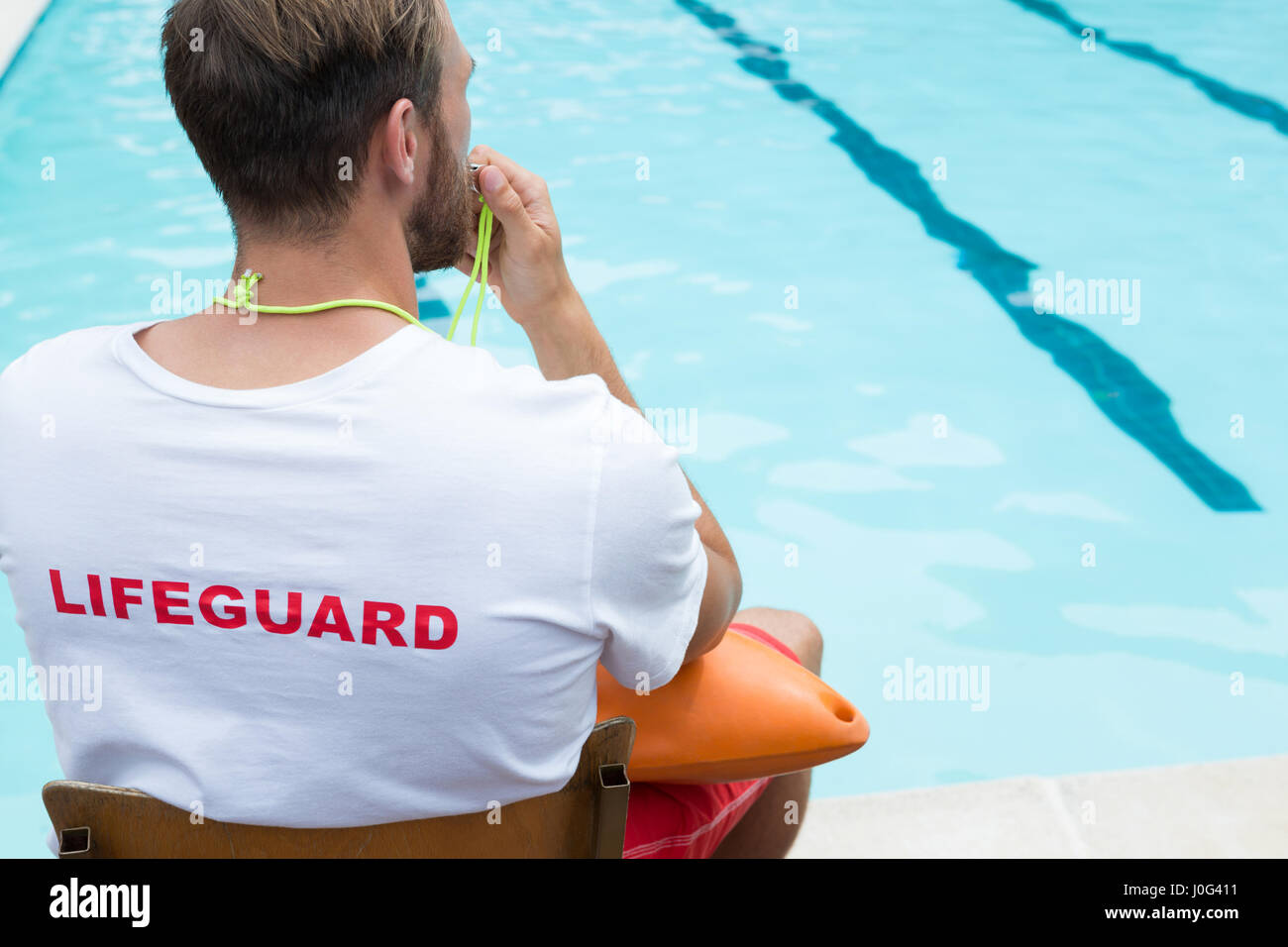 Rear view of lifeguard sitting on chair and blowing whistle at poolside ...