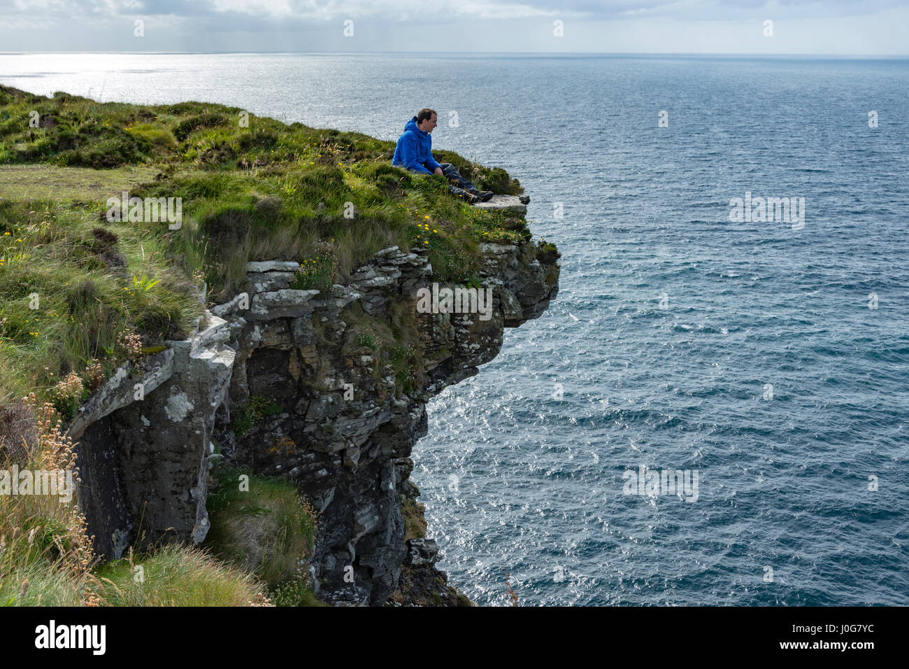 A person sat on an overhanging cliff edge at the Cliffs of Moher ...