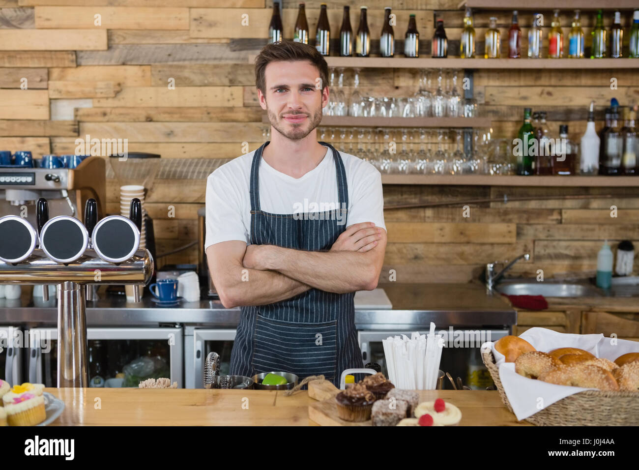 Portrait of smiling waiter standing with arms crossed at counter in ...