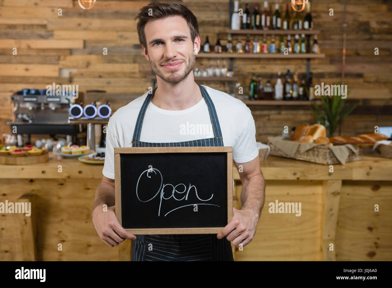 Portrait of smiling waiter standing with open signboard at counter in ...