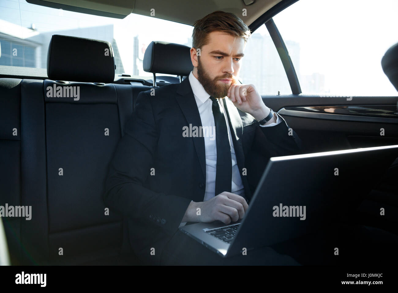 Serious business man using laptop computer while sitting on back seat ...