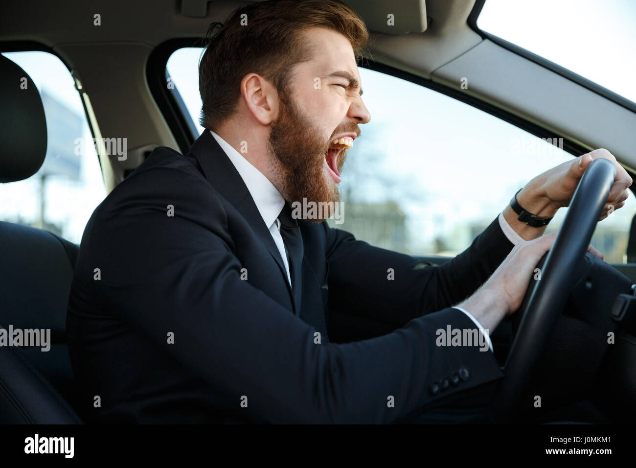 Side view of screaming bearded business man in suit driving a car and ...