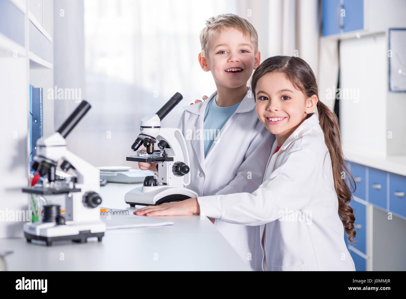 Little boy and girl in white coats using microscope and smiling at ...