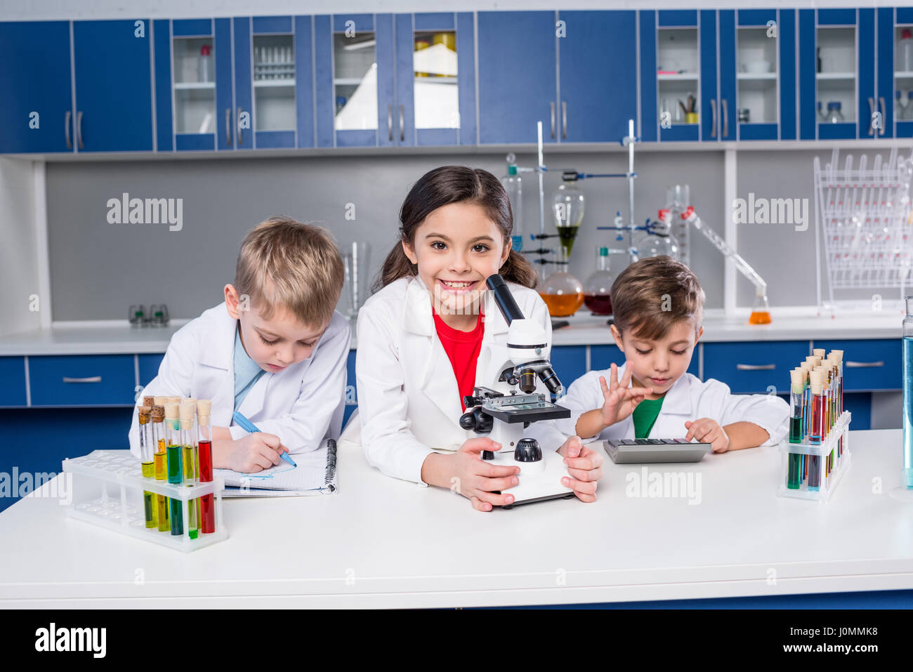 Three kids in chemical laboratory making notes and using microscope and ...