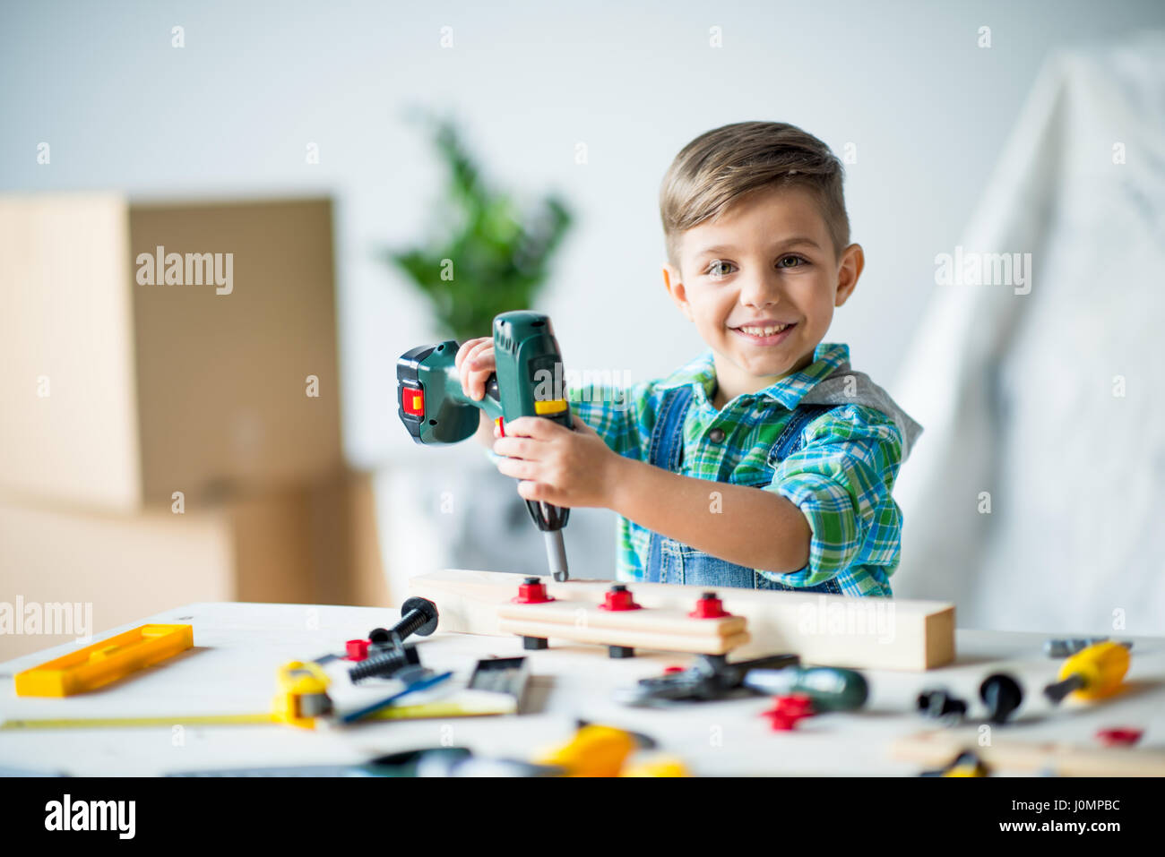 Smiling little boy drilling wooden plank with electric drill at table ...
