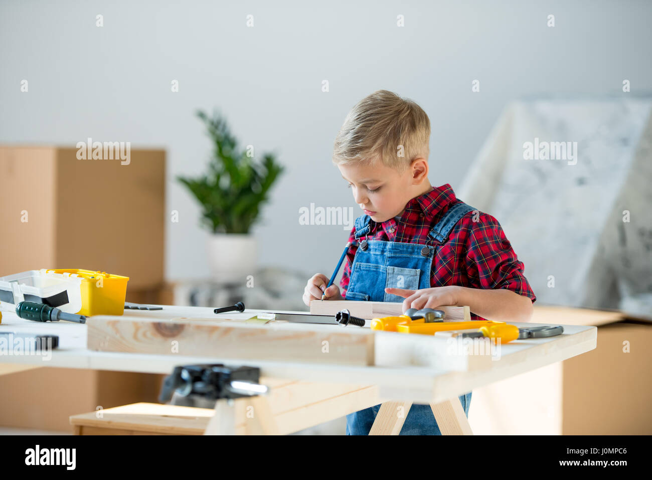 Pensive little boy standing at table with toy tools and marking wooden ...
