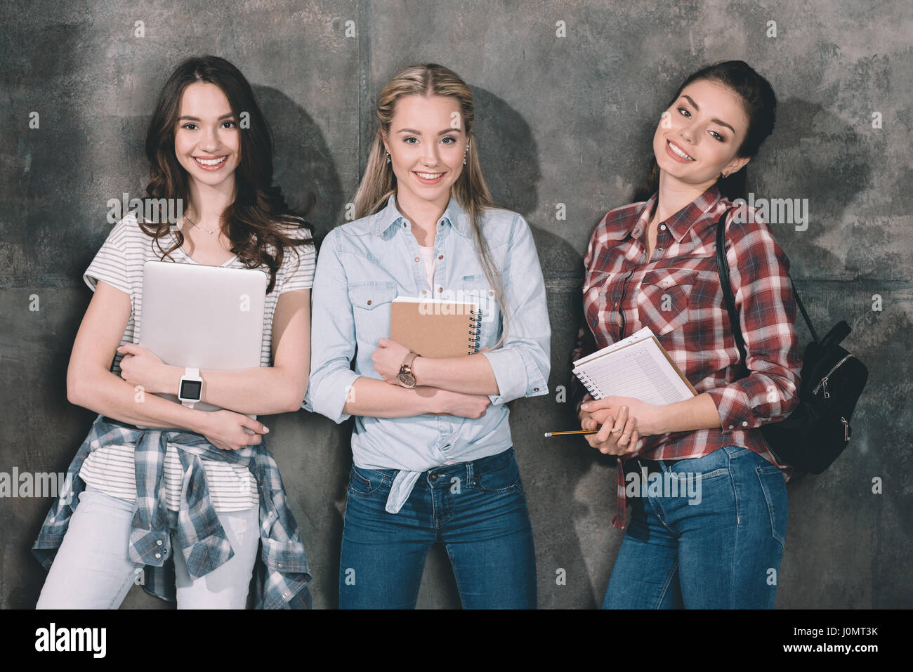 Three attractive students with copybooks and laptop standing at wall ...