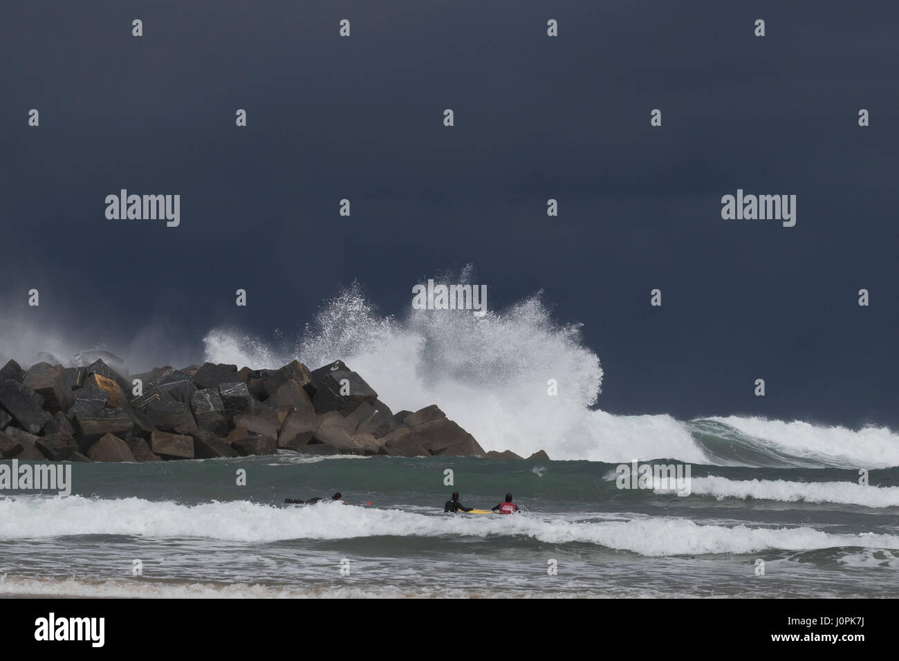 Surfers in the water observing a big wave breaking in breakwater blocks ...