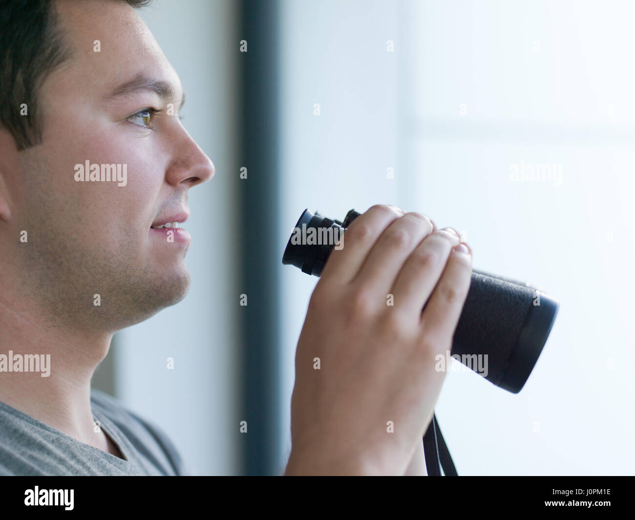 Young man standing looking through a glass window with binoculars as he ...