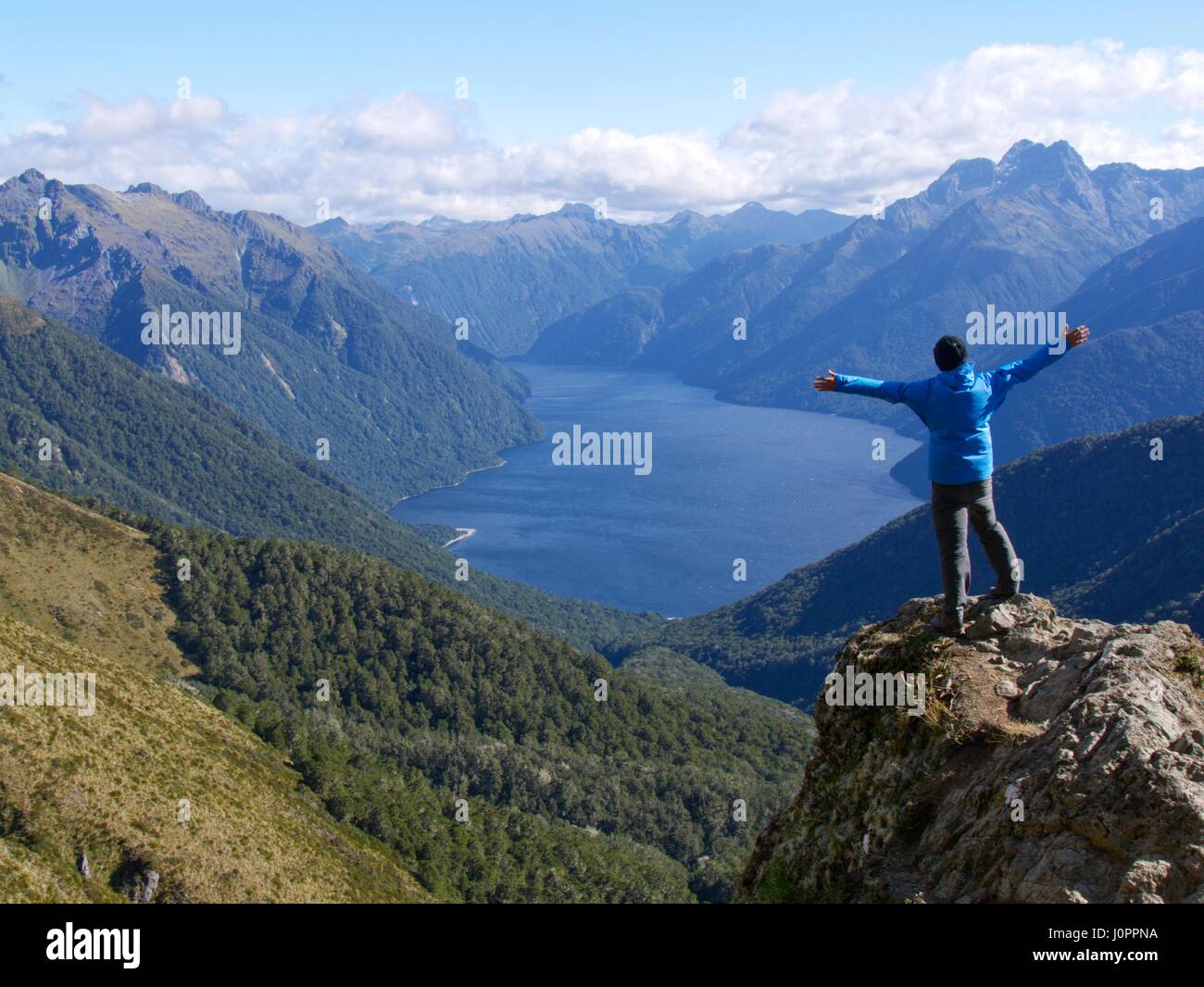Young man in blue jacket standing on mountain ridge with lifted arms ...