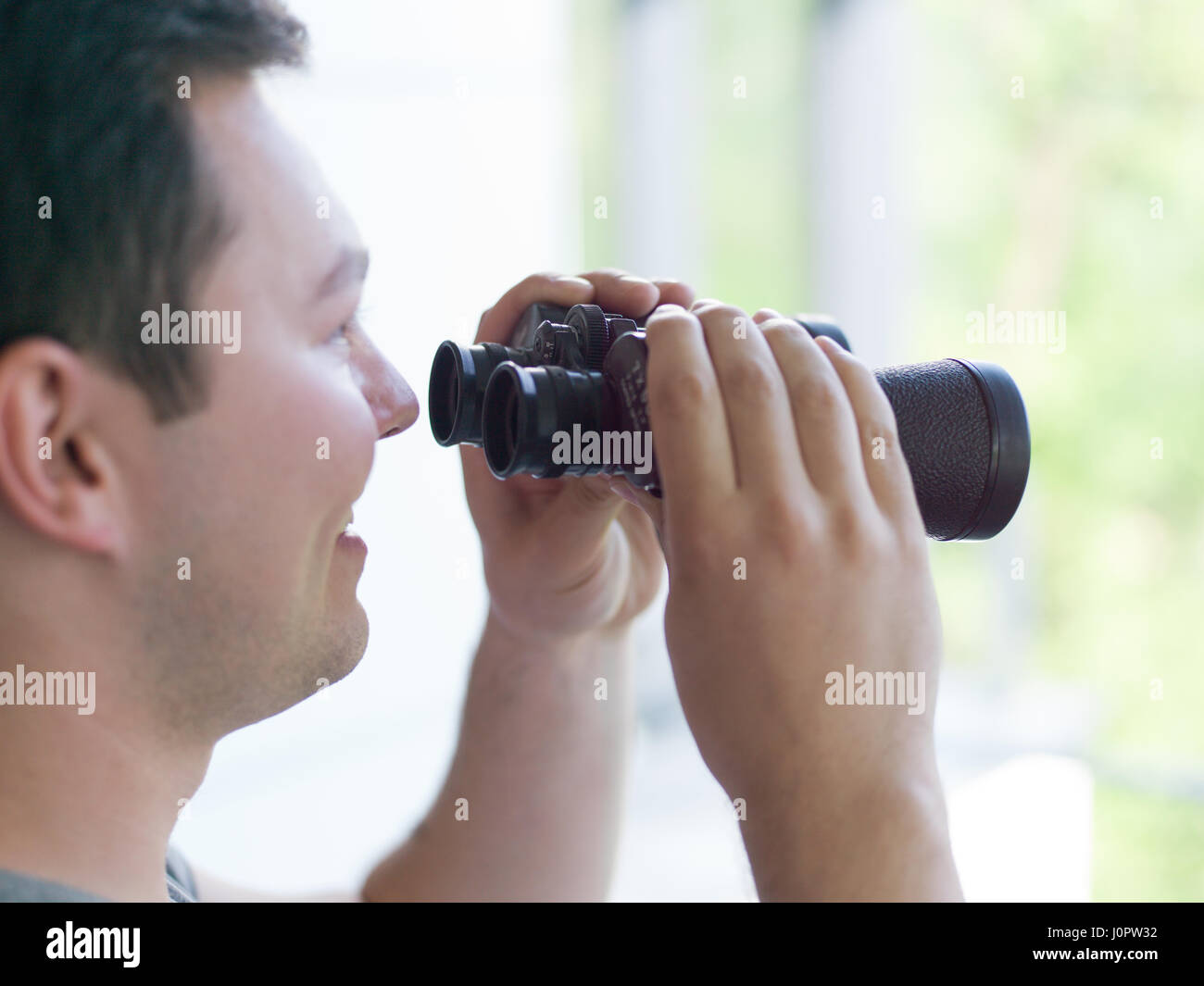 Young man standing looking through a glass window with binoculars as he ...