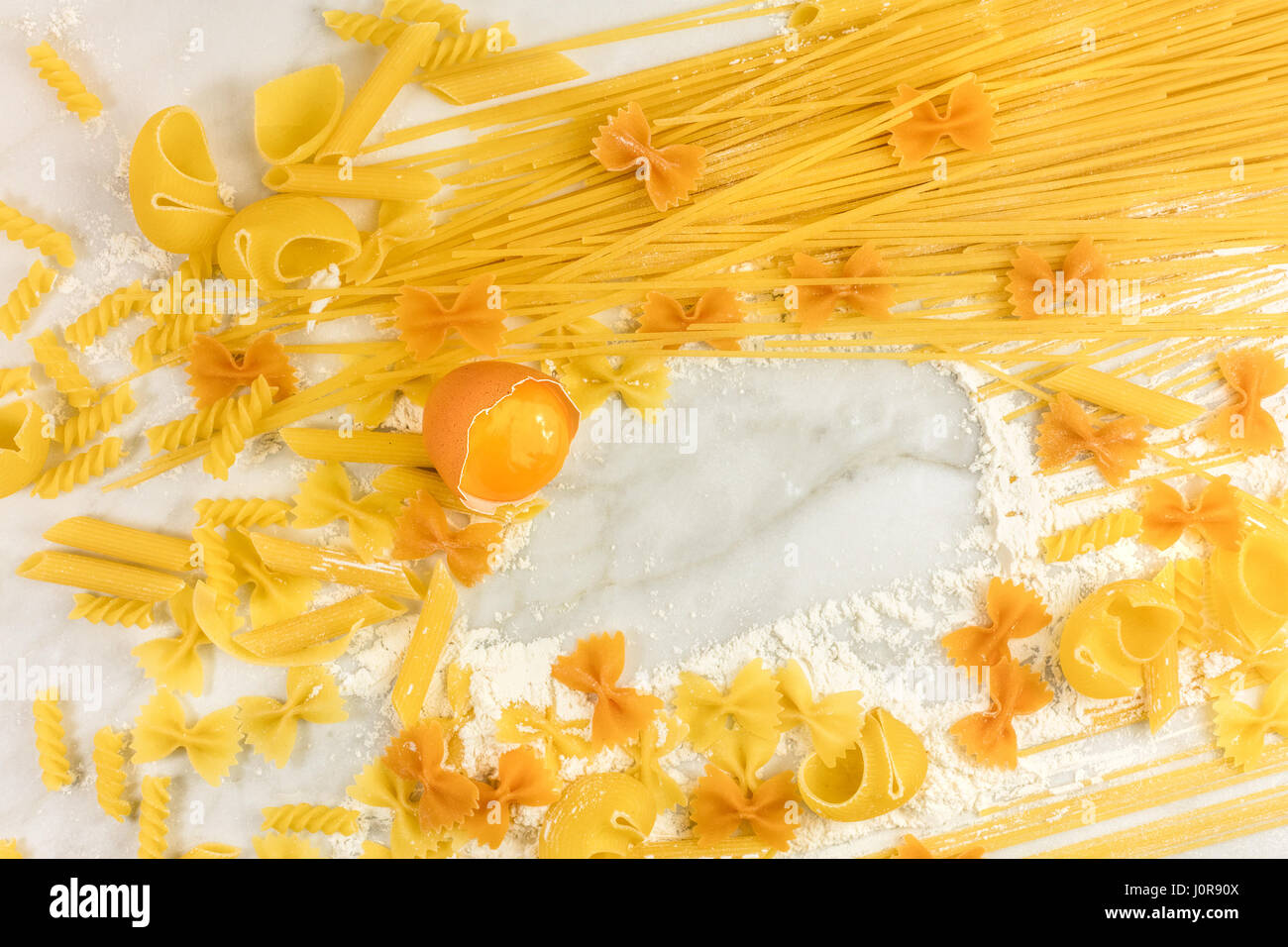 Various types of pasta on a white marble table with flour and an egg ...