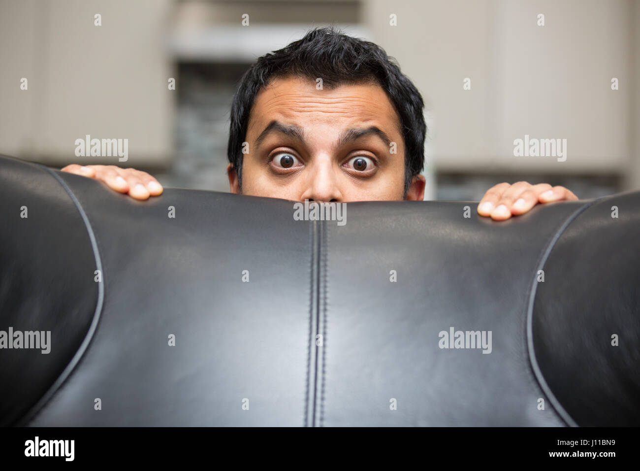 Closeup portrait, grown young man hiding behind black leather couch ...