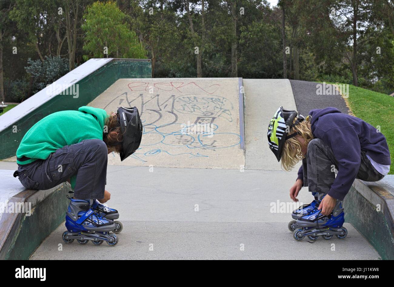 Two boys sitting on a skate ramp doing up their inline skates or roller ...
