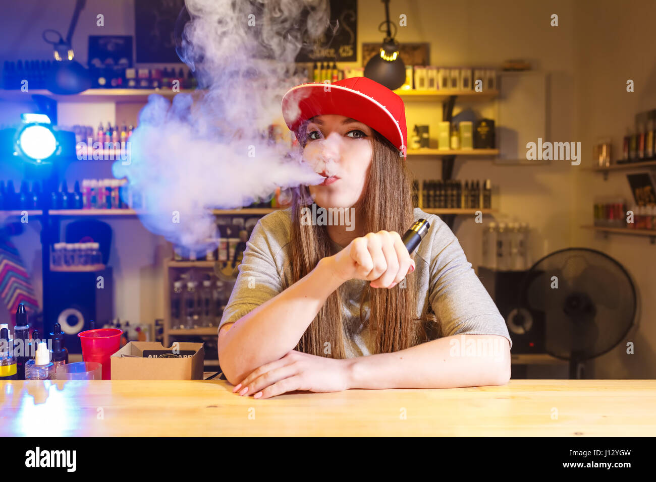 Young pretty woman in red cap smoke an electronic cigarette at the vape ...