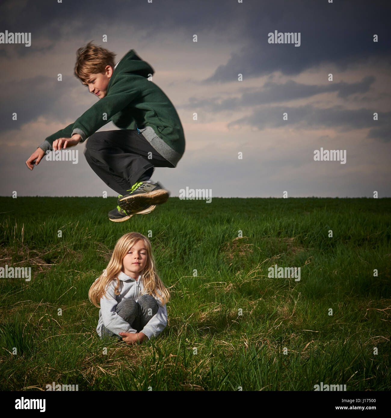 Brother jumping over sister in meadow, cloudy sky background. Children ...