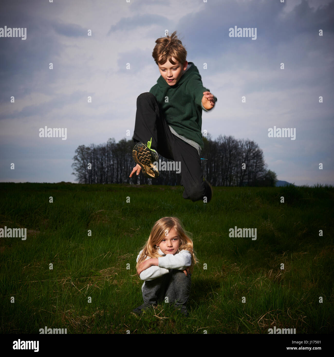 Brother jumping over sister in meadow, cloudy sky background. Children ...