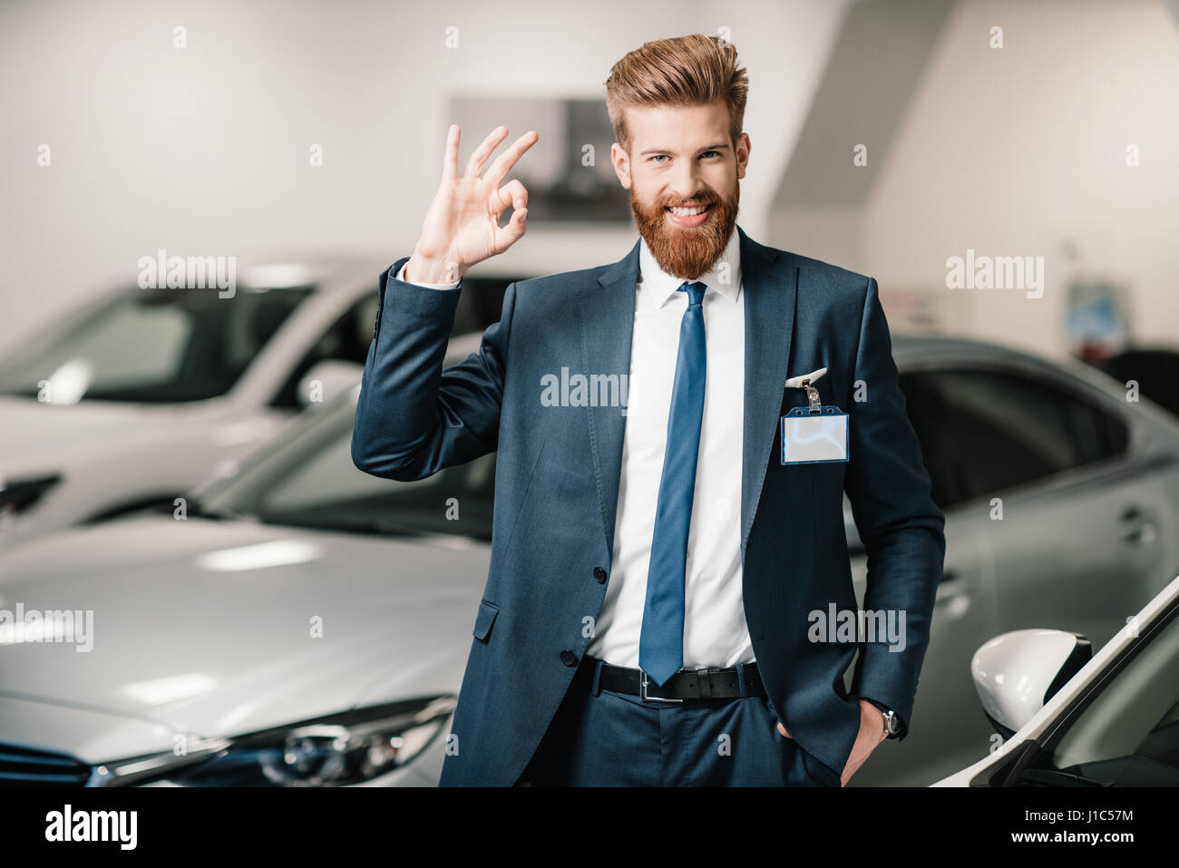 salesman in suit showing ok sign and looking at camera in dealership ...