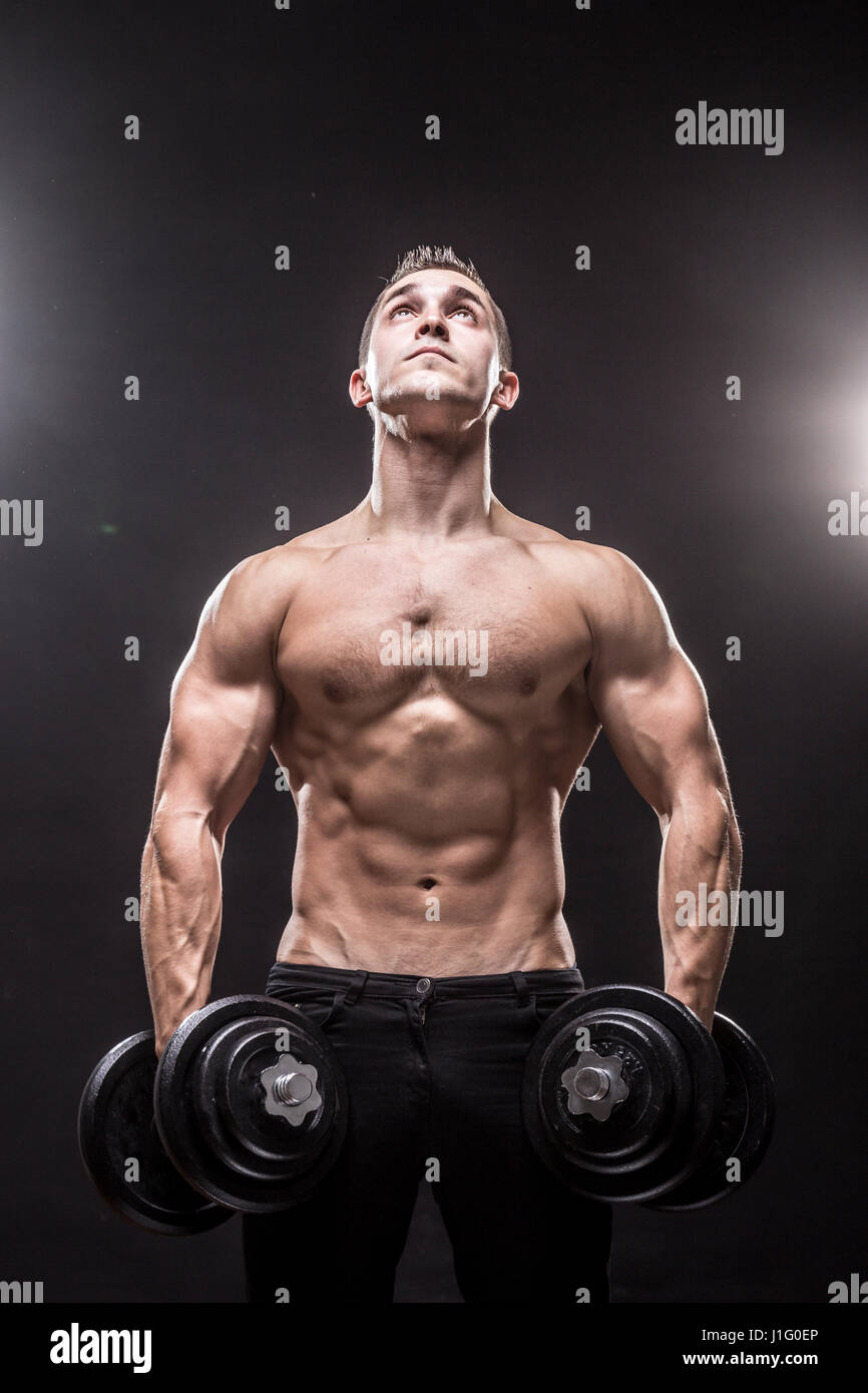 young man bodybuilder looking up, holding dumbbells. studio reflectors ...