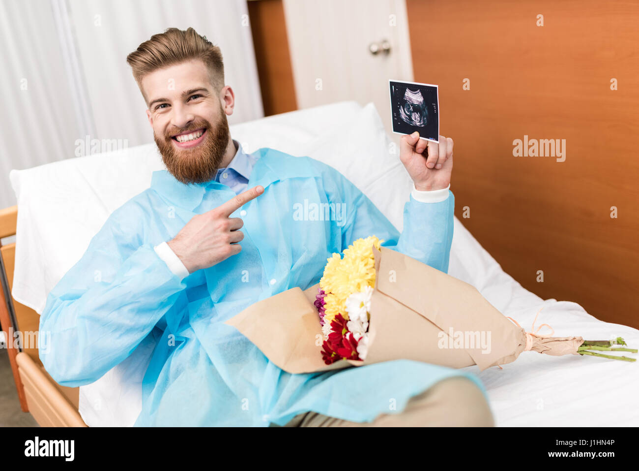 front view of happy young man with flowers and sonogram in hospital ...