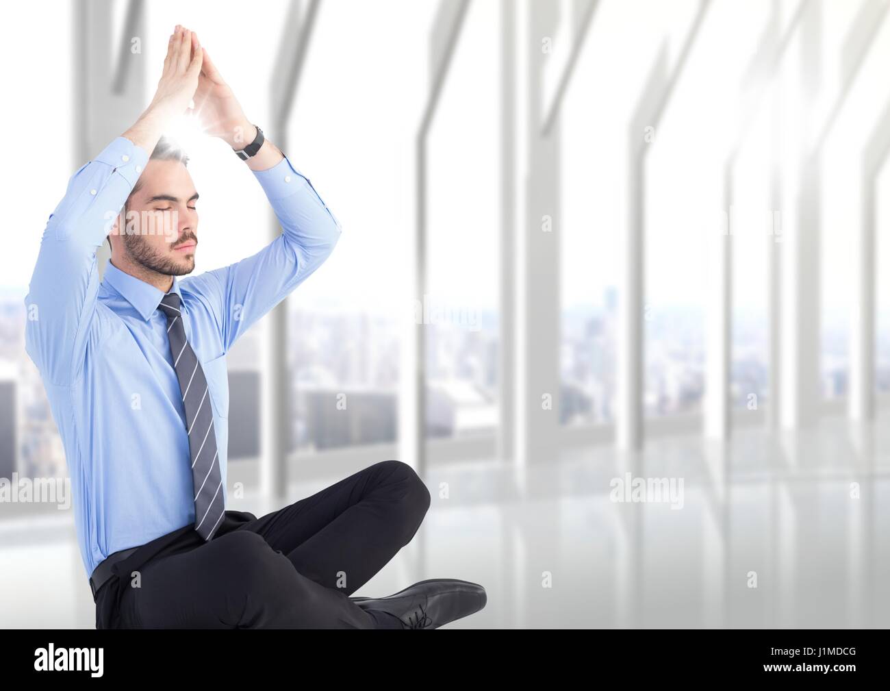 Business man with hands over head meditating with flare against blurry ...