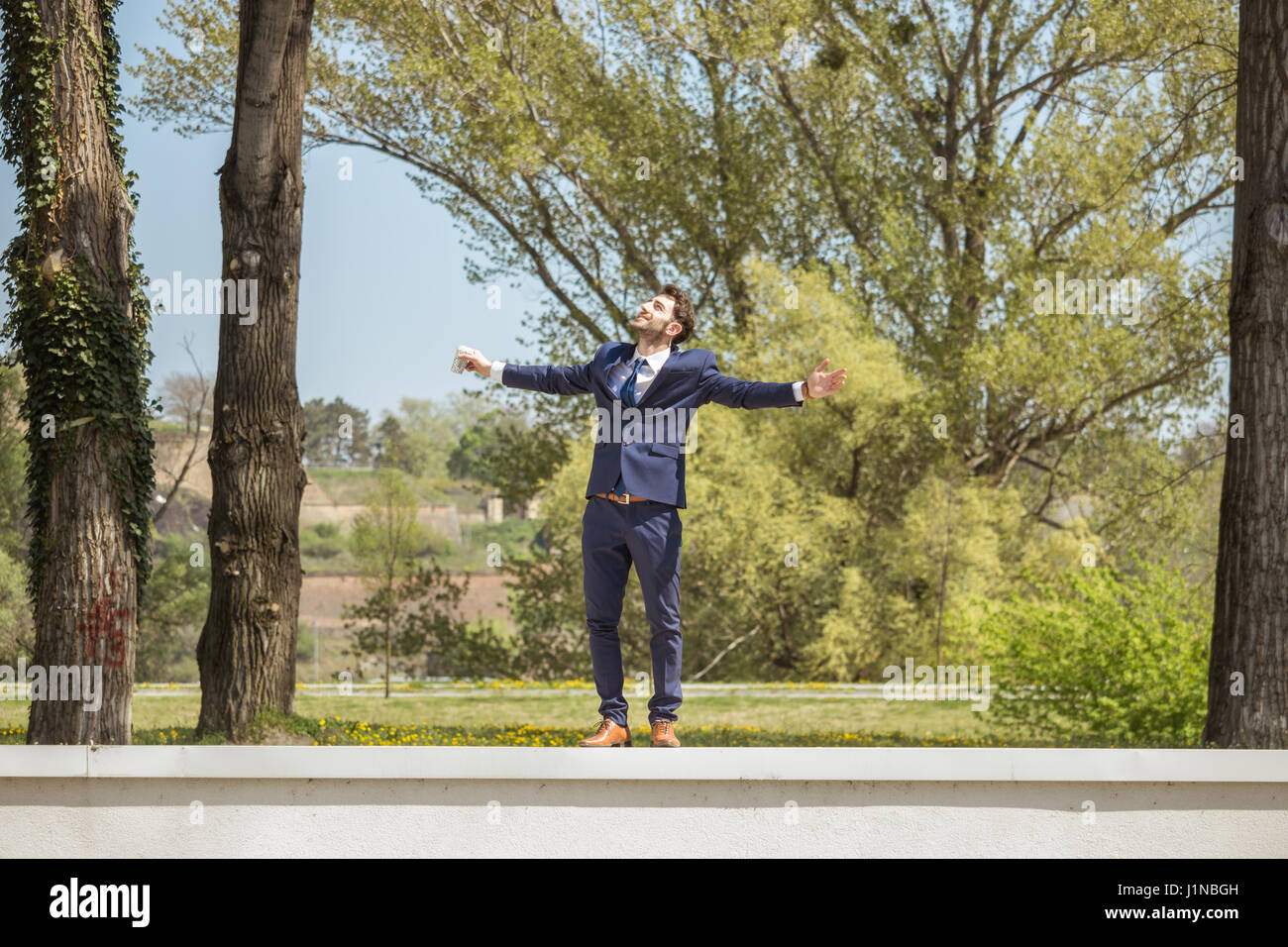 young man, looking up above, standing on wall, arms outstreched, sunny ...