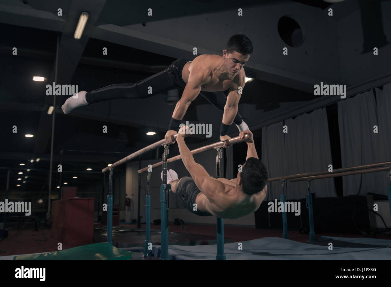 Two young adult men, gymnast in air, parallel bars. Indoors, dark hall ...