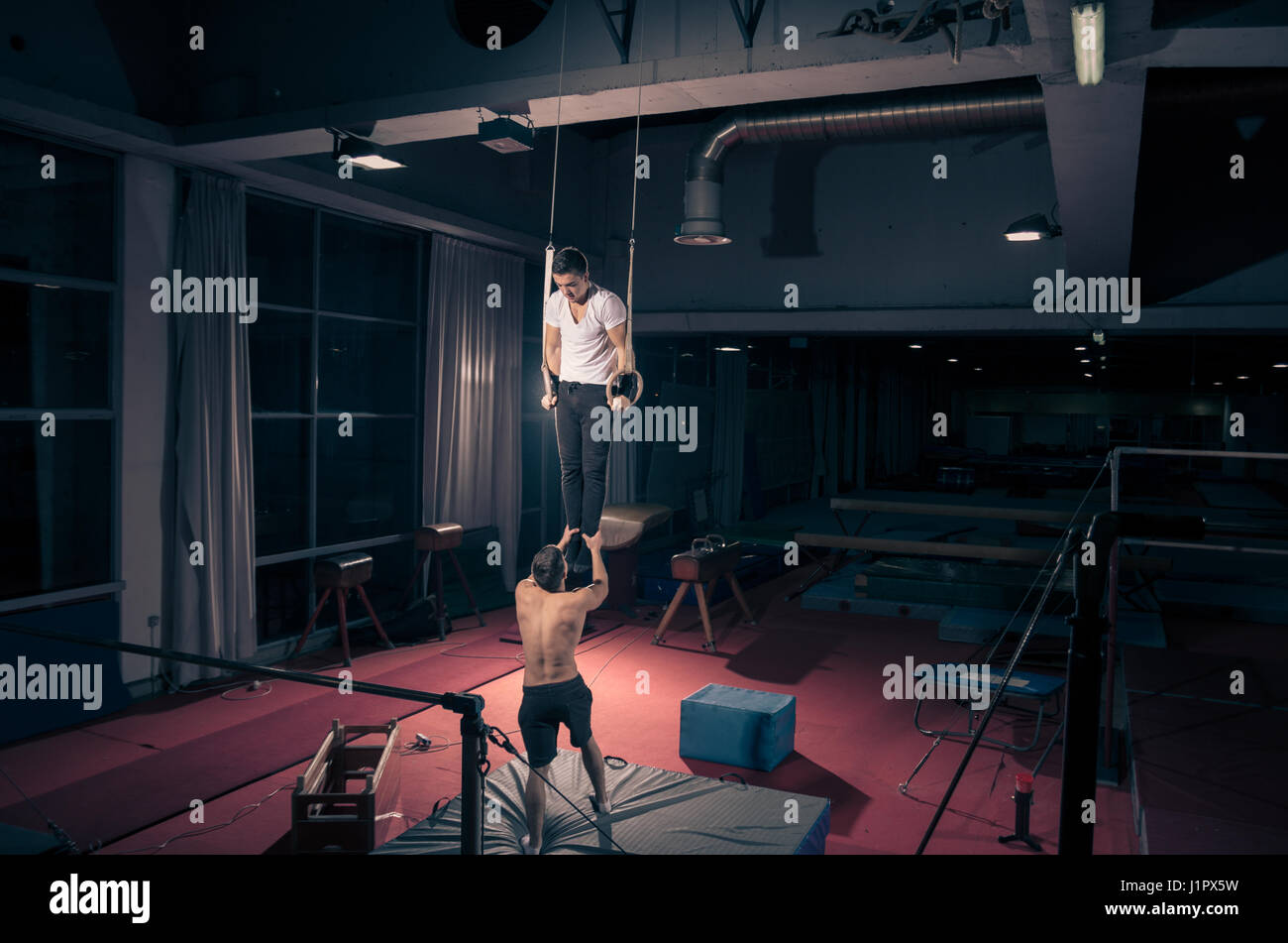 Two men gymnasts holding helping, gymnastics rings. Indoors, dark hall ...