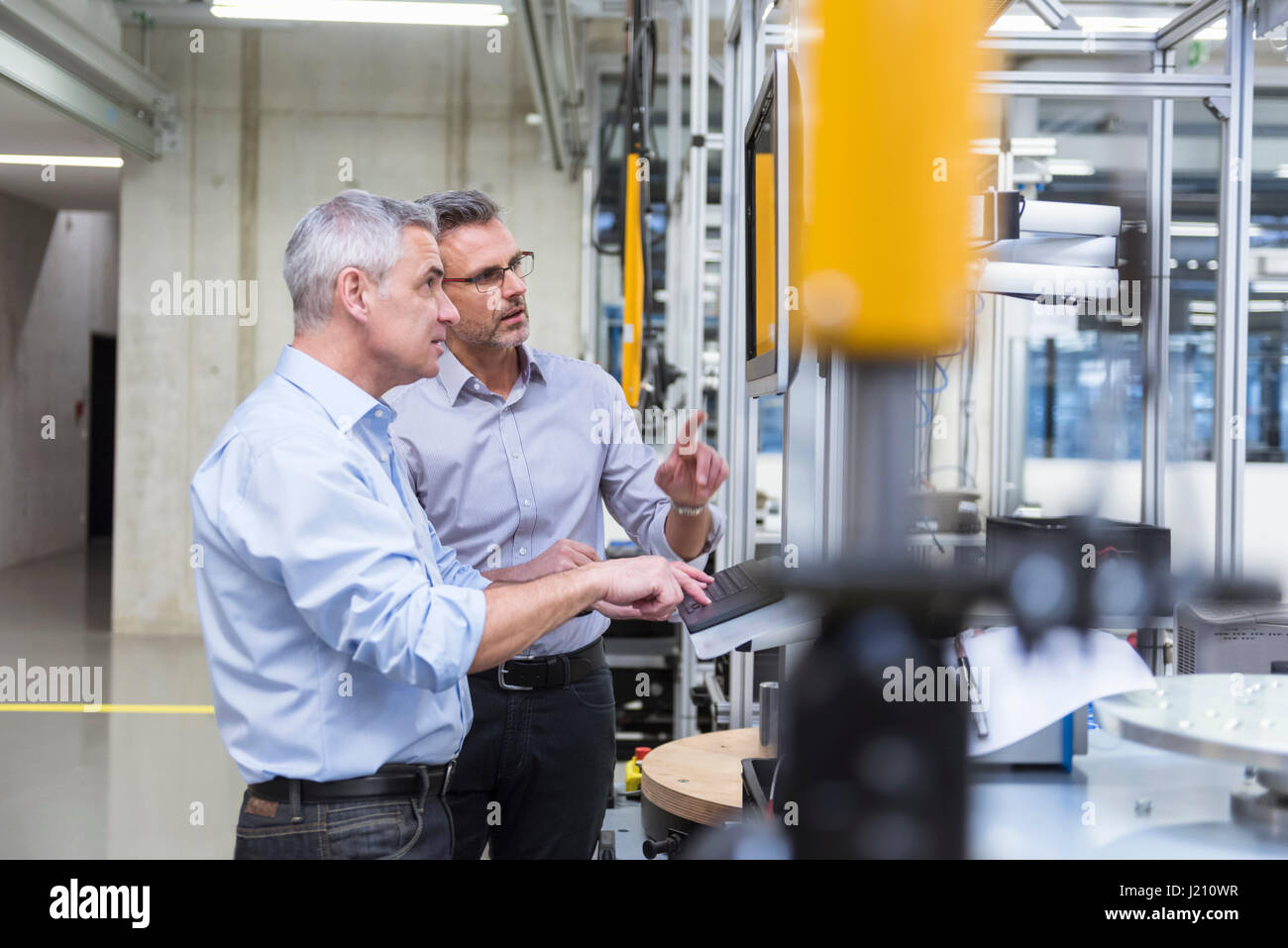 Two businessmen in modern factory hall talking at computer monitor ...