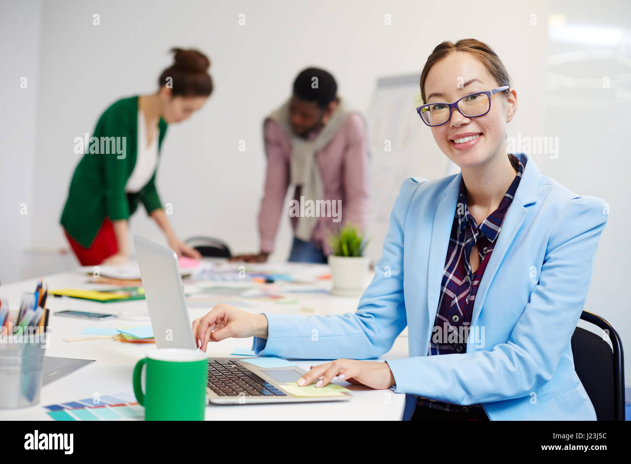 Attractive young woman with laptop networking in office on background ...