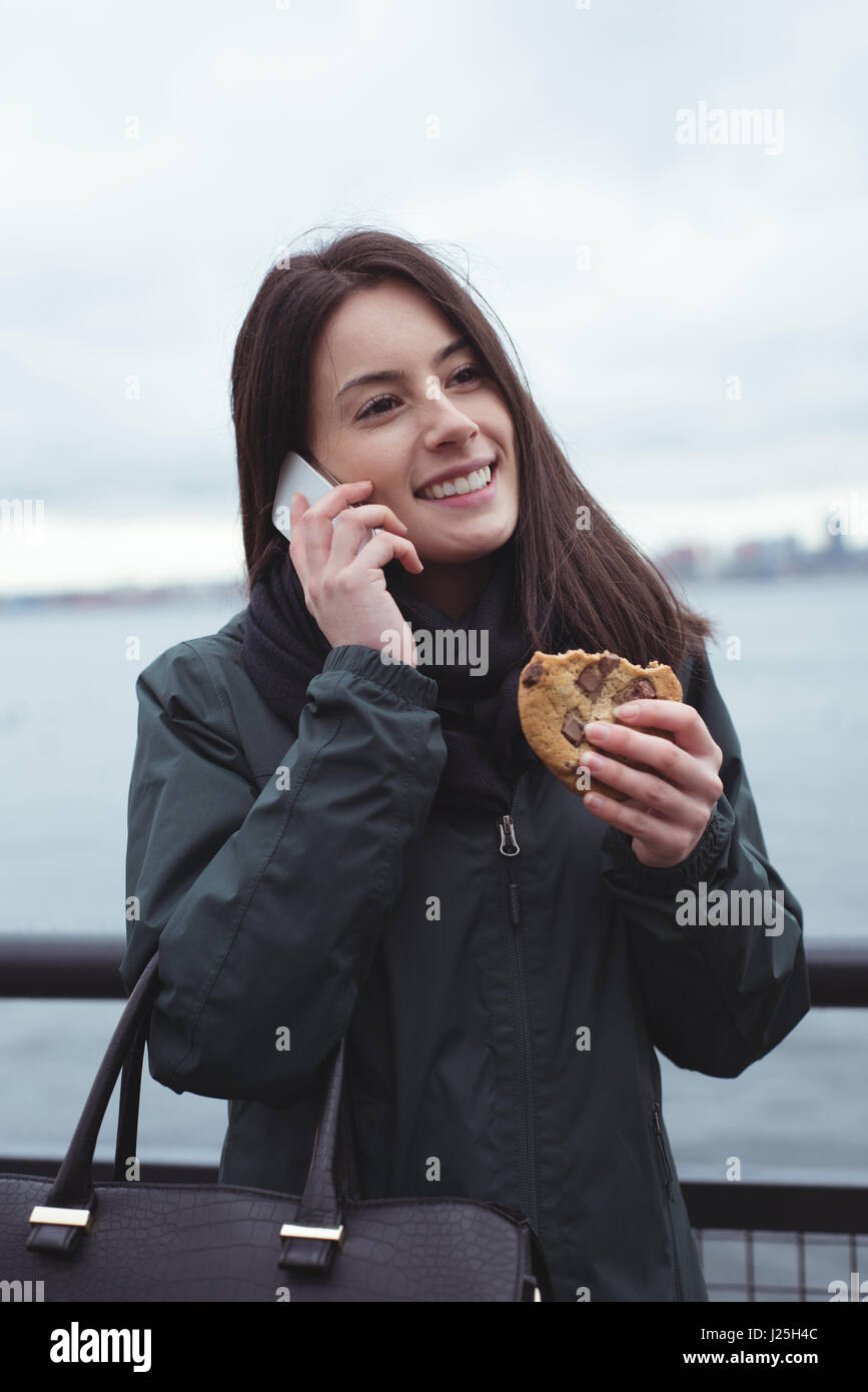 Happy woman talking on phone while eating food against railing in city ...