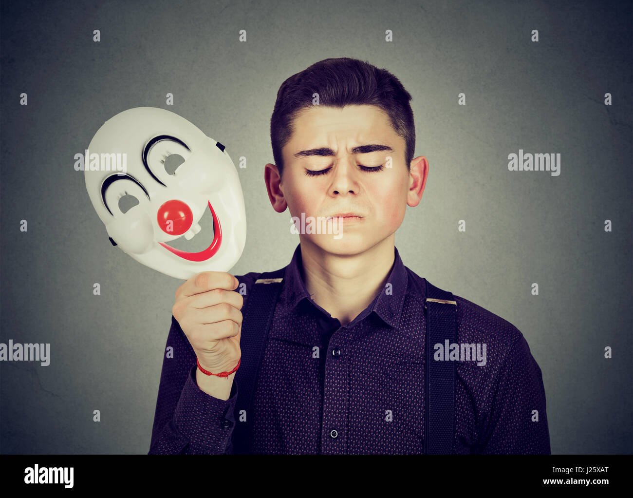 Young sad man taking off happy clown mask isolated on gray wall ...