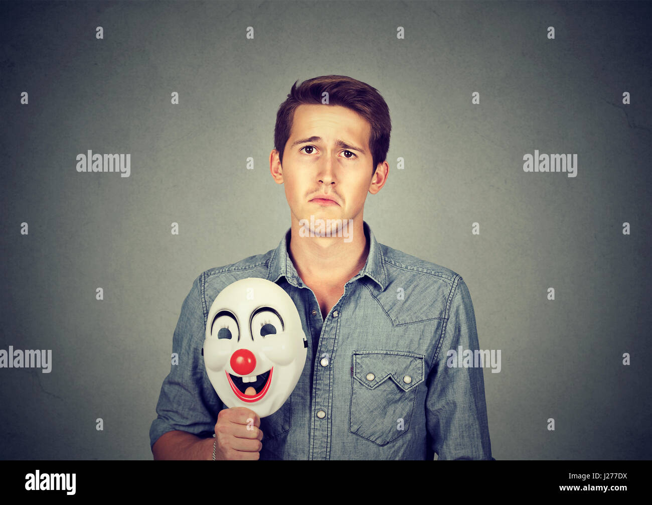 Young sad man with happy clown mask isolated on gray wall background ...