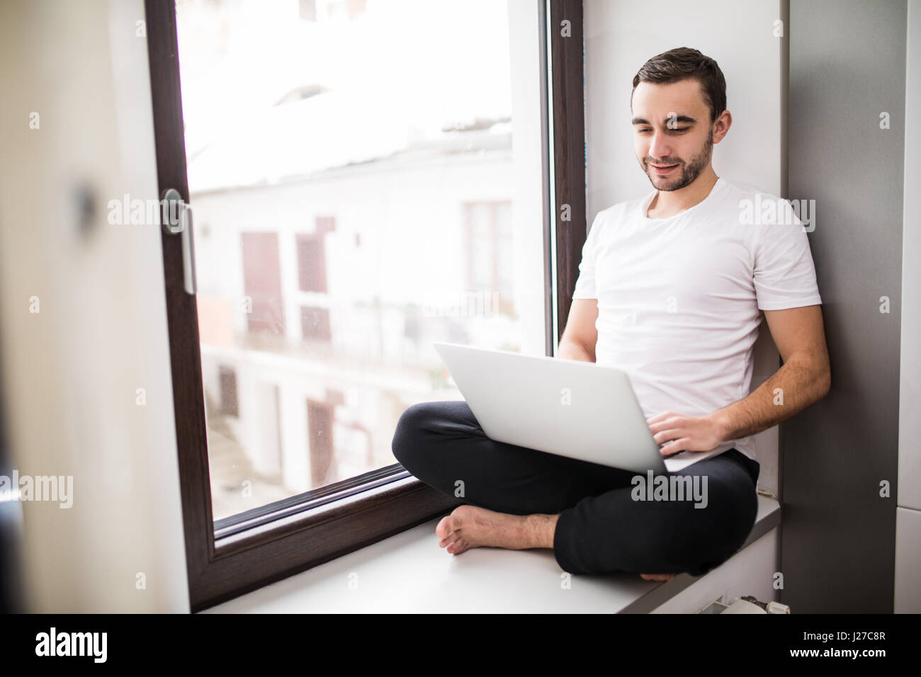 Attractive man using a laptop, looking at camera and smiling while ...